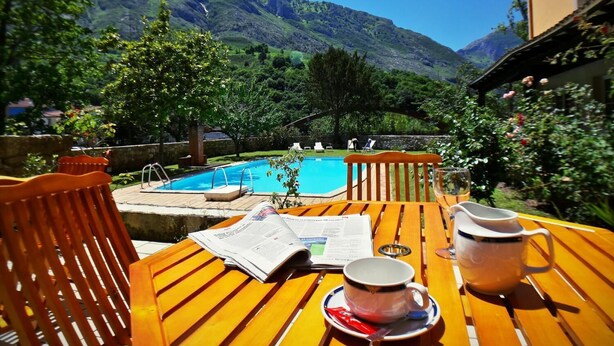 Uma mesa de madeira com jornal, uma xícara e um copo, com vista para uma piscina e montanhas ao fundo.