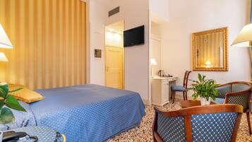 Quarto de hotel com cama, escrivaninha, cadeira, televisão, espelho e planta.