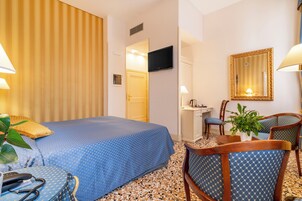 Quarto de hotel com cama, escrivaninha, cadeira, televisão, espelho e planta.