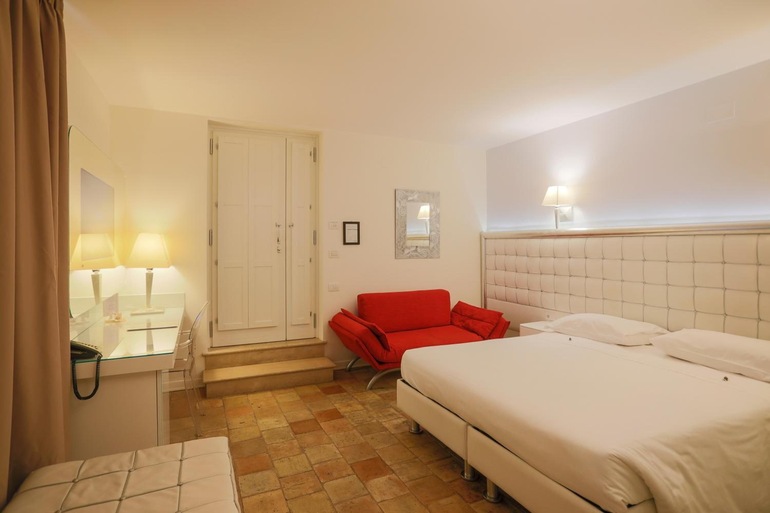 Quarto de hotel com uma cama, uma cadeira vermelha, uma escrivaninha com abajur e um guarda-roupa.