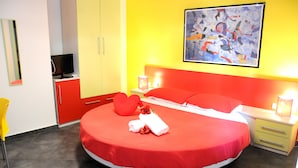 Uma cama redonda com roupa de cama vermelha, um travesseiro em forma de coração vermelho e uma toalha branca com uma rosa vermelha. Há uma pequena mesa de cabeceira com um abajur, uma cadeira amarela e uma TV sobre um suporte.