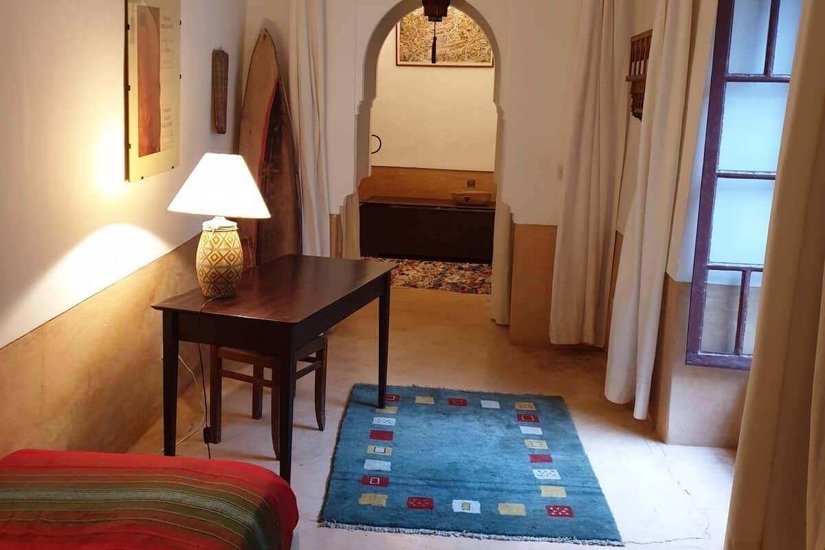 Quarto com cama, escrivaninha com abajur, tapete e porta arqueada que dá para outro cômodo.