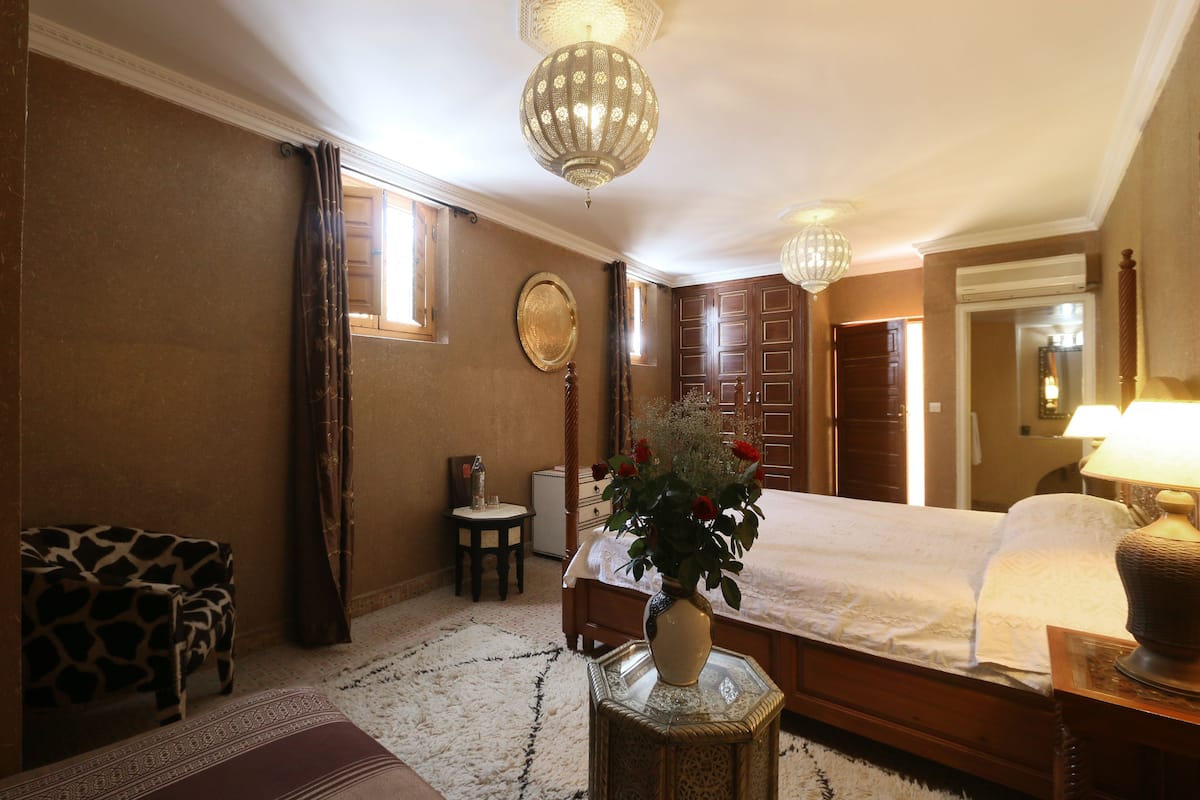 Quarto de hotel com cama, uma pequena mesa com um vaso de flores, uma cadeira, espelho redondo e um lustre.