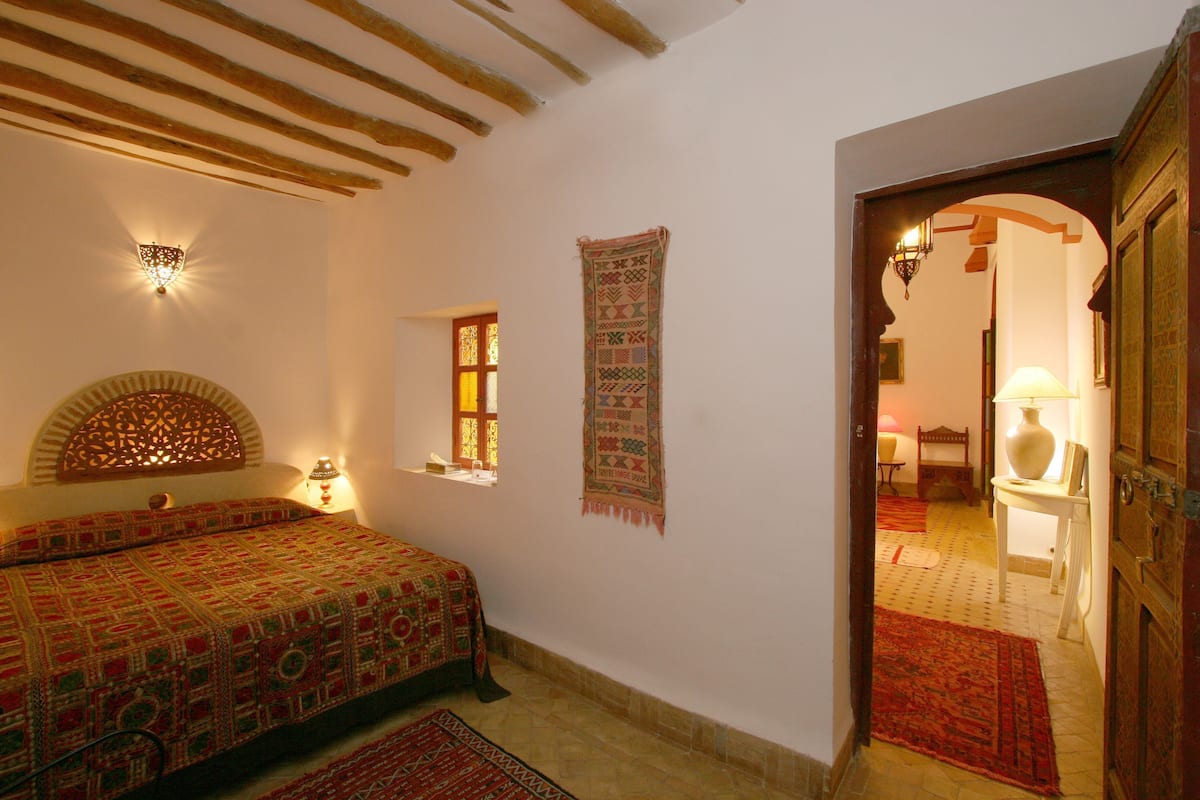 Quarto com cama, janela, um objeto decorativo na parede e porta que dá para outro cômodo.