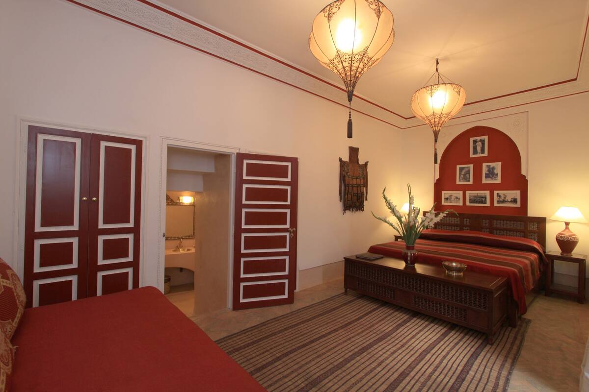 Quarto de hotel com uma cama, uma secretária, duas luminárias suspensas, uma parede vermelha com quadros e uma porta que dá para outro quarto.