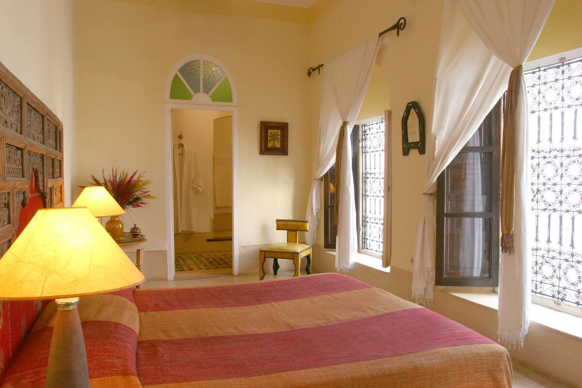 Quarto de hotel com uma cama, duas luminárias, uma cadeira e uma janela com cortinas.