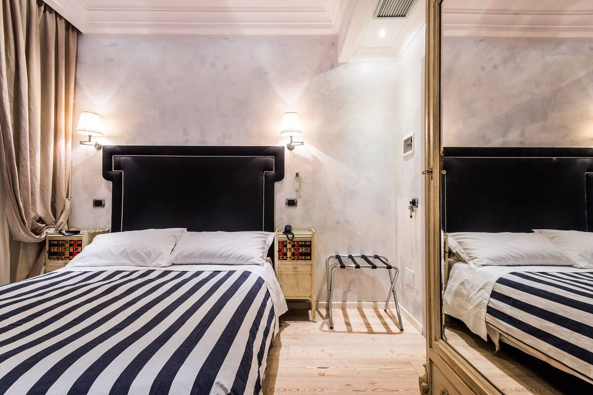 Quarto de hotel com duas camas, uma mesinha de cabeceira, um espelho e piso de madeira.