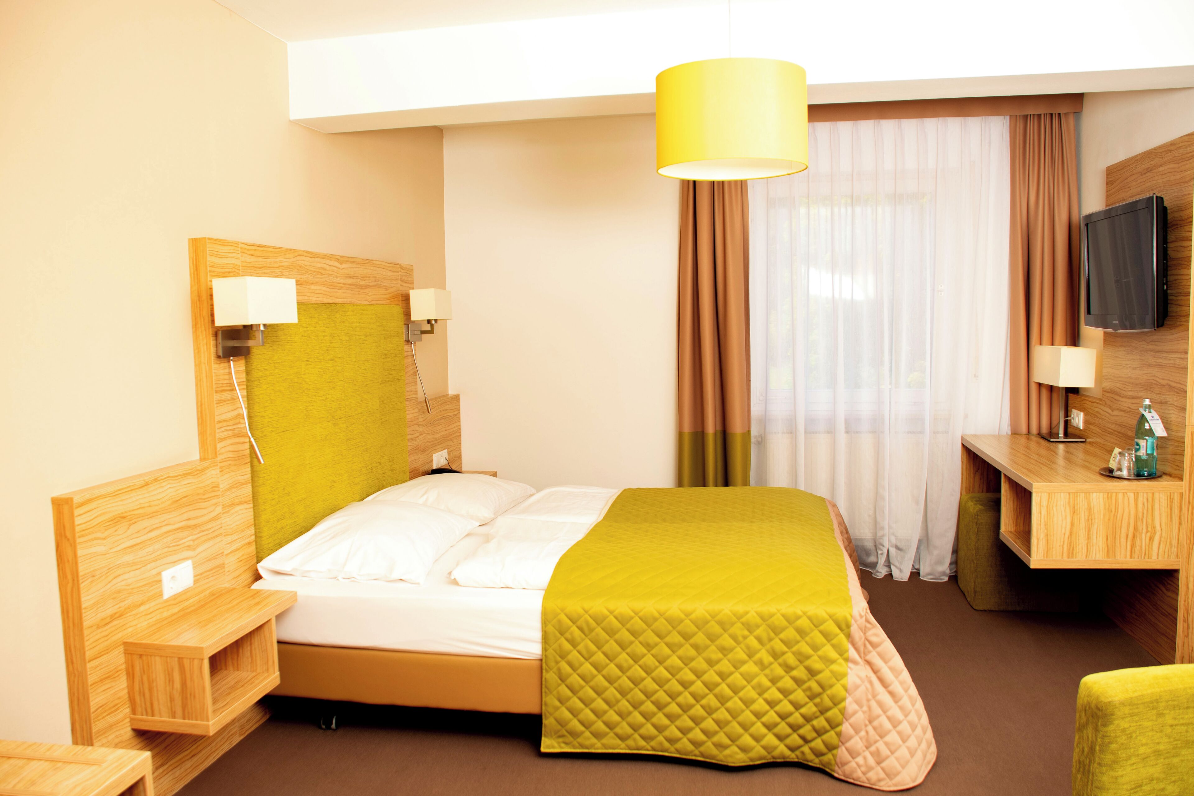 Quarto de hotel com cama, mesas de cabeceira, escrivaninha com cadeira, televisão e janela com cortinas.