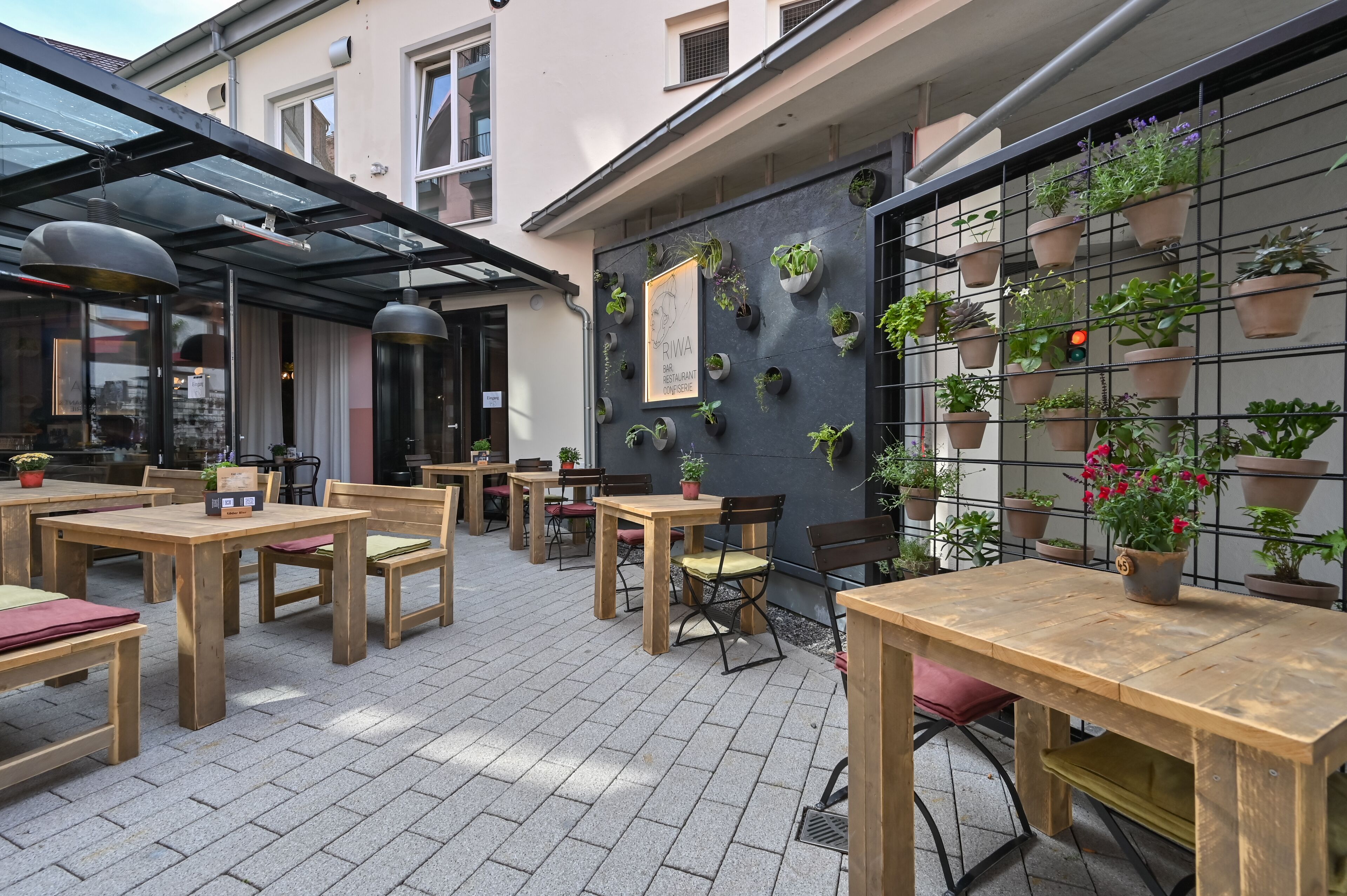 Uma área de café ao ar livre com mesas e cadeiras de madeira, plantas em vasos numa parede e um corrimão de metal.
