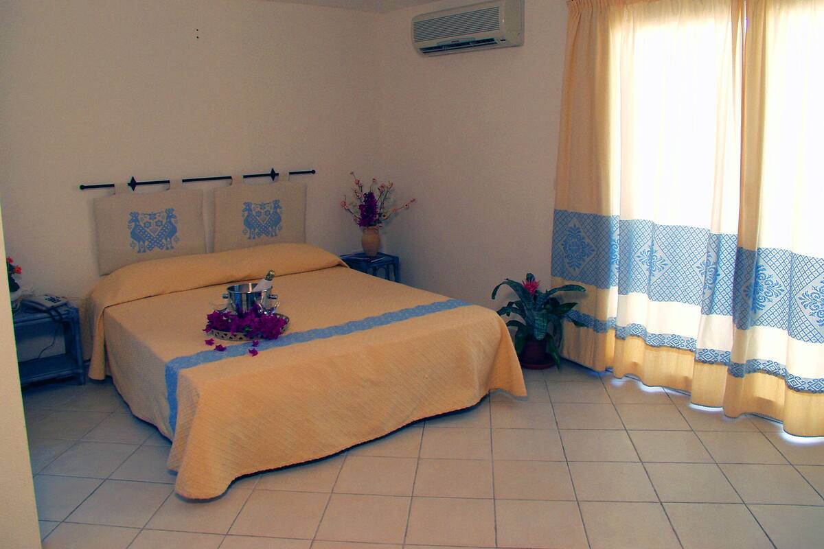 Quarto com cama, mesa de cabeceira, ar condicionado, janela com cortinas e um vaso de planta.