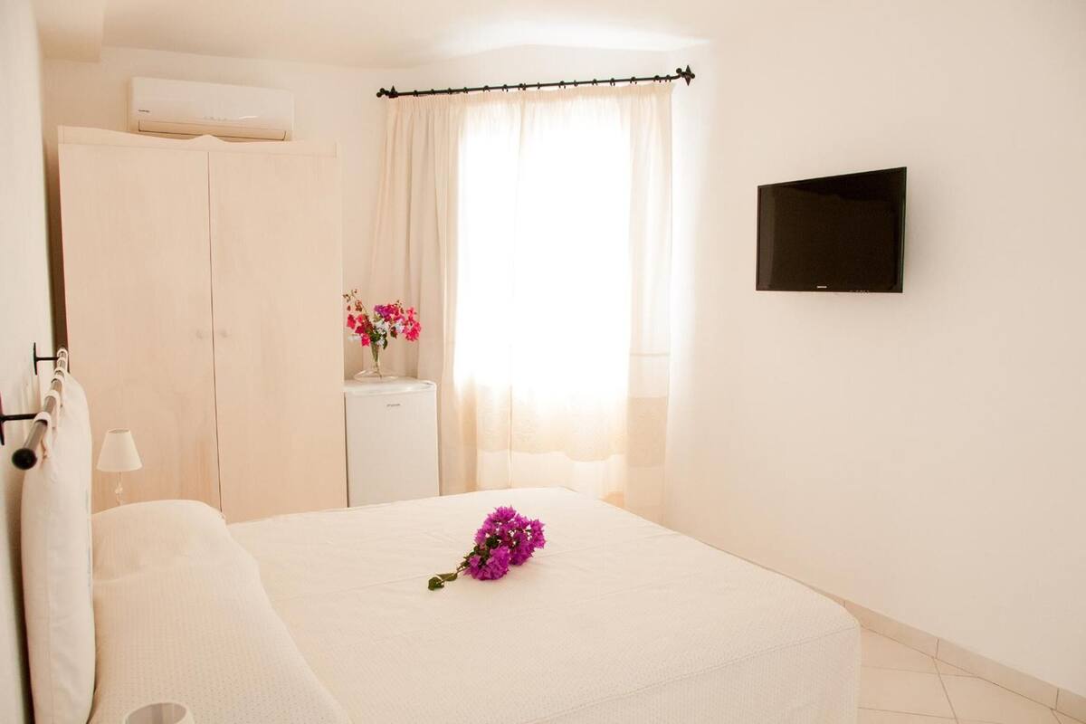 Quarto de hotel com uma cama, televisão na parede, guarda-roupa e uma mesinha com um vaso de flores.