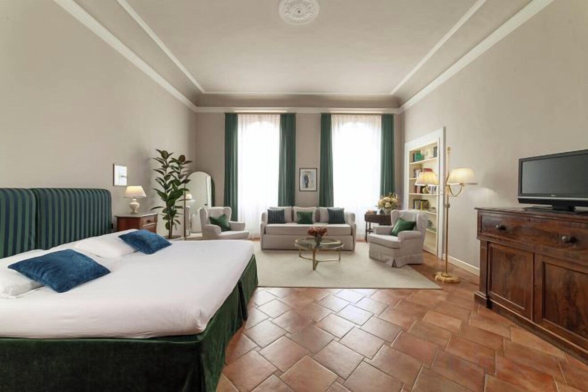 Quarto de hotel com cama, duas poltronas, um sofá, uma mesa de centro, uma televisão e uma estante de livros.