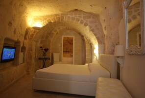 Um quarto com arco de pedra, uma cama, mesa de cabeceira com um vaso e uma televisão na parede.
