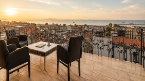 Terraço com mesa e cadeiras, vista para a paisagem urbana e o mar ao pôr do sol.