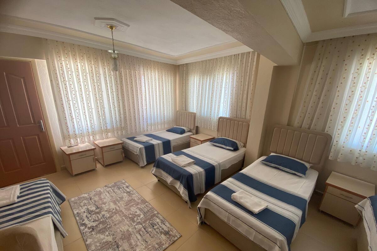 Quarto de hotel com duas camas de solteiro, uma secretária pequena, uma cadeira, mesa de cabeceira e uma janela grande com cortinas.