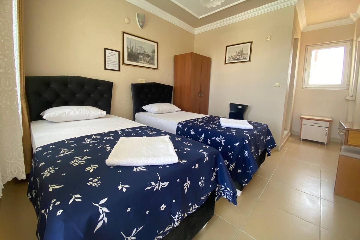 Quarto de hotel com duas camas, um guarda-roupa, uma pequena mesa de trabalho e uma janela.
