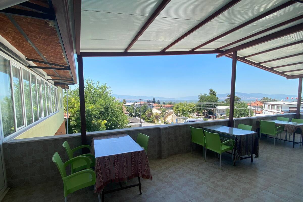 Uma área de estar coberta ao ar livre, com mesas e cadeiras, vista para uma cidade e montanhas ao longe, e chão de azulejos.