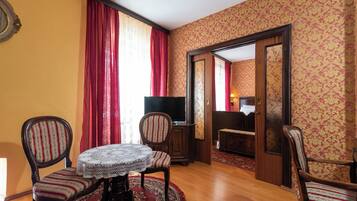 Quarto com piso de madeira, uma mesa redonda com toalha de renda, duas cadeiras listradas, uma televisão e um espelho refletindo um quarto com cortina vermelha e papel de parede estampado.