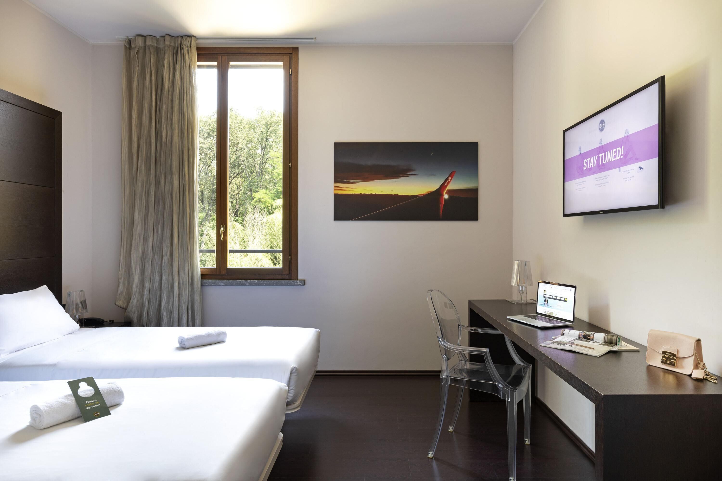 Quarto de hotel com duas camas, uma escrivaninha com um laptop, uma televisão e uma janela com cortinas.