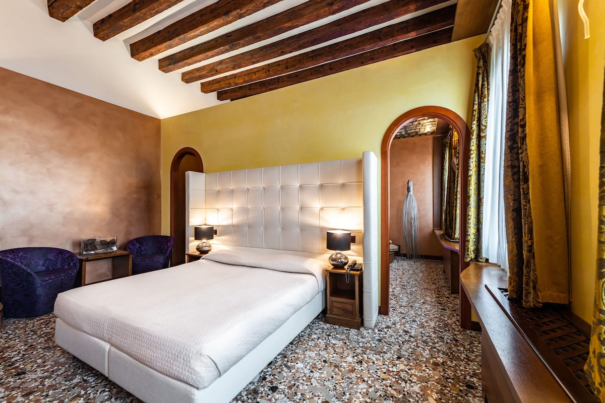 Quarto de hotel com uma cama grande, duas mesas de cabeceira com luminárias, uma mesinha com um vaso e um banco junto à parede.