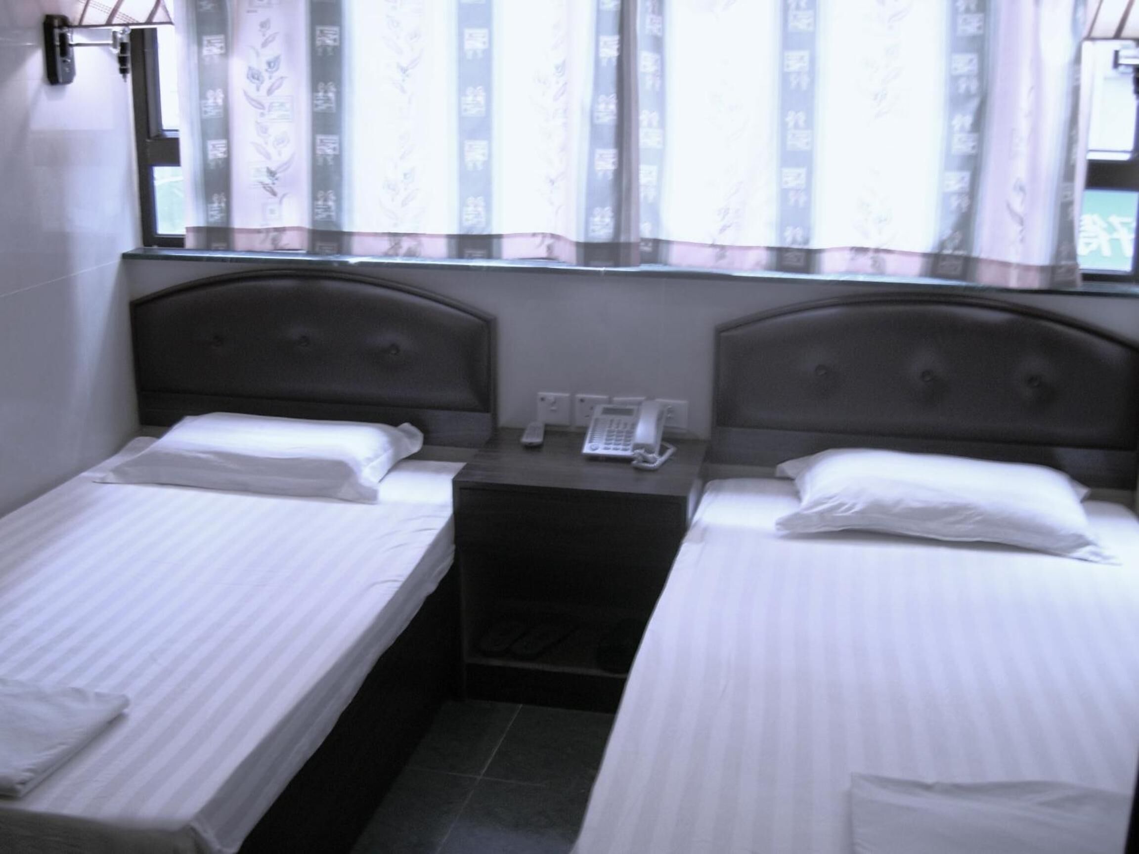 Quarto de hotel com duas camas de solteiro, uma mesinha com telefone e uma janela com cortinas.