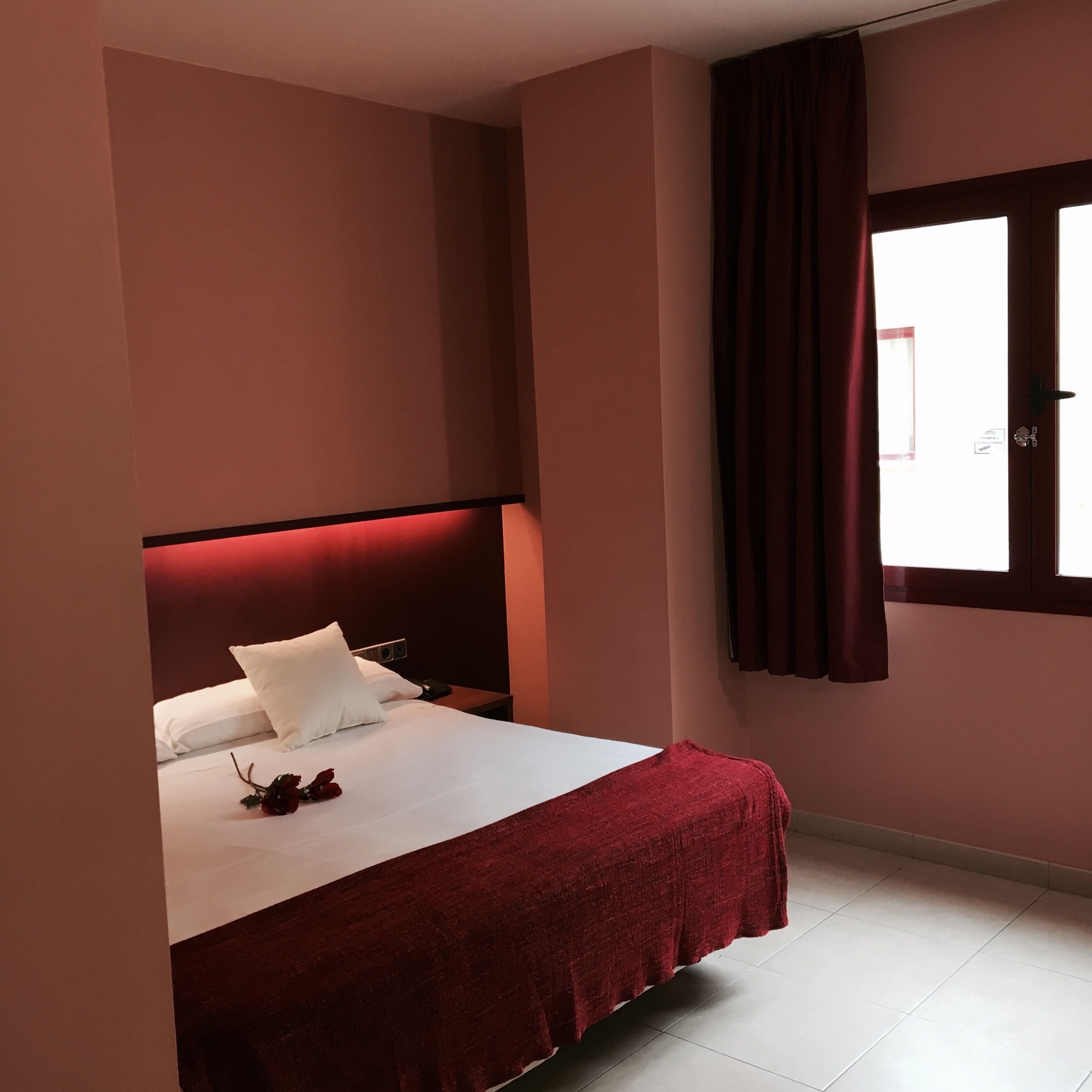Quarto com cama, cabeceira, janela com cortinas e piso de azulejos.