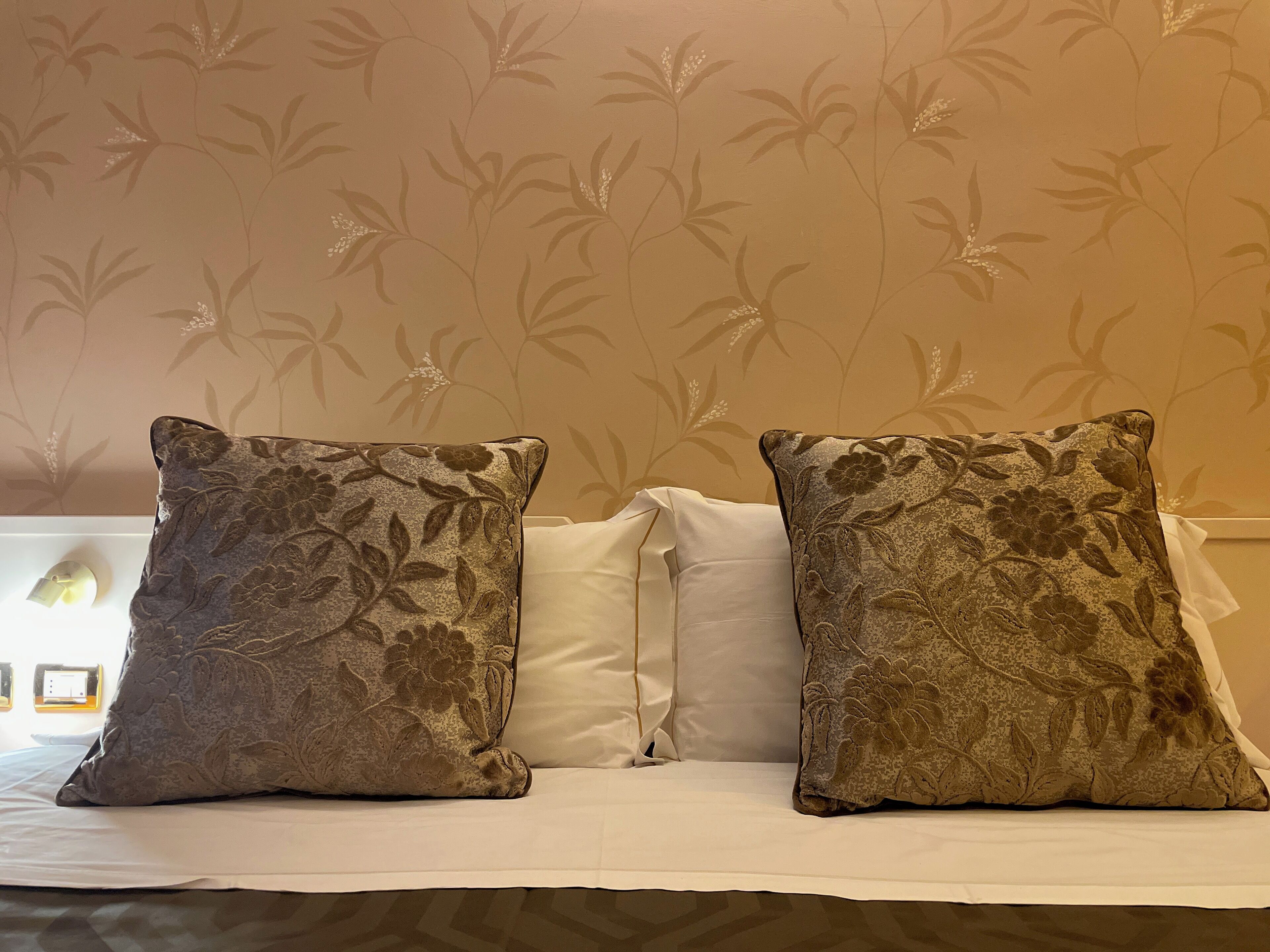 Uma cama com duas almofadas decorativas e uma almofada branca simples, apoiada em uma parede com padrão floral.