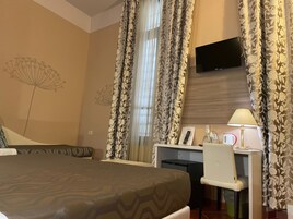 Quarto de hotel com cama, escrivaninha, cadeira, televisão e cortinas.