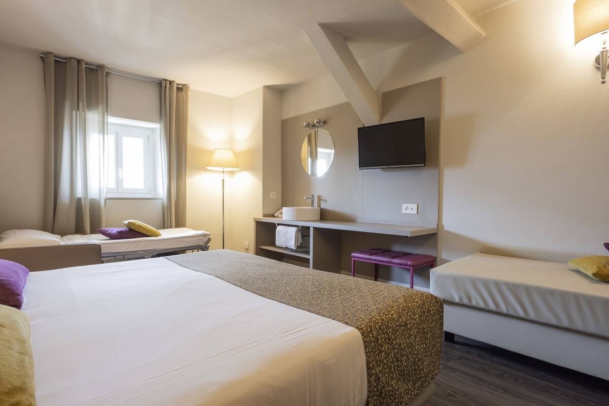 Quarto de hotel com uma cama grande, uma escrivaninha com cadeira, televisão fixada na parede, janela com cortinas e um banquinho pequeno.