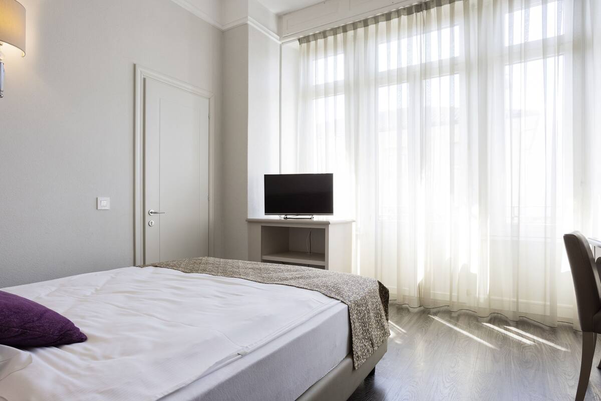 Quarto de hotel com uma cama, televisão sobre um suporte, uma cadeira e uma janela com cortinas translúcidas.