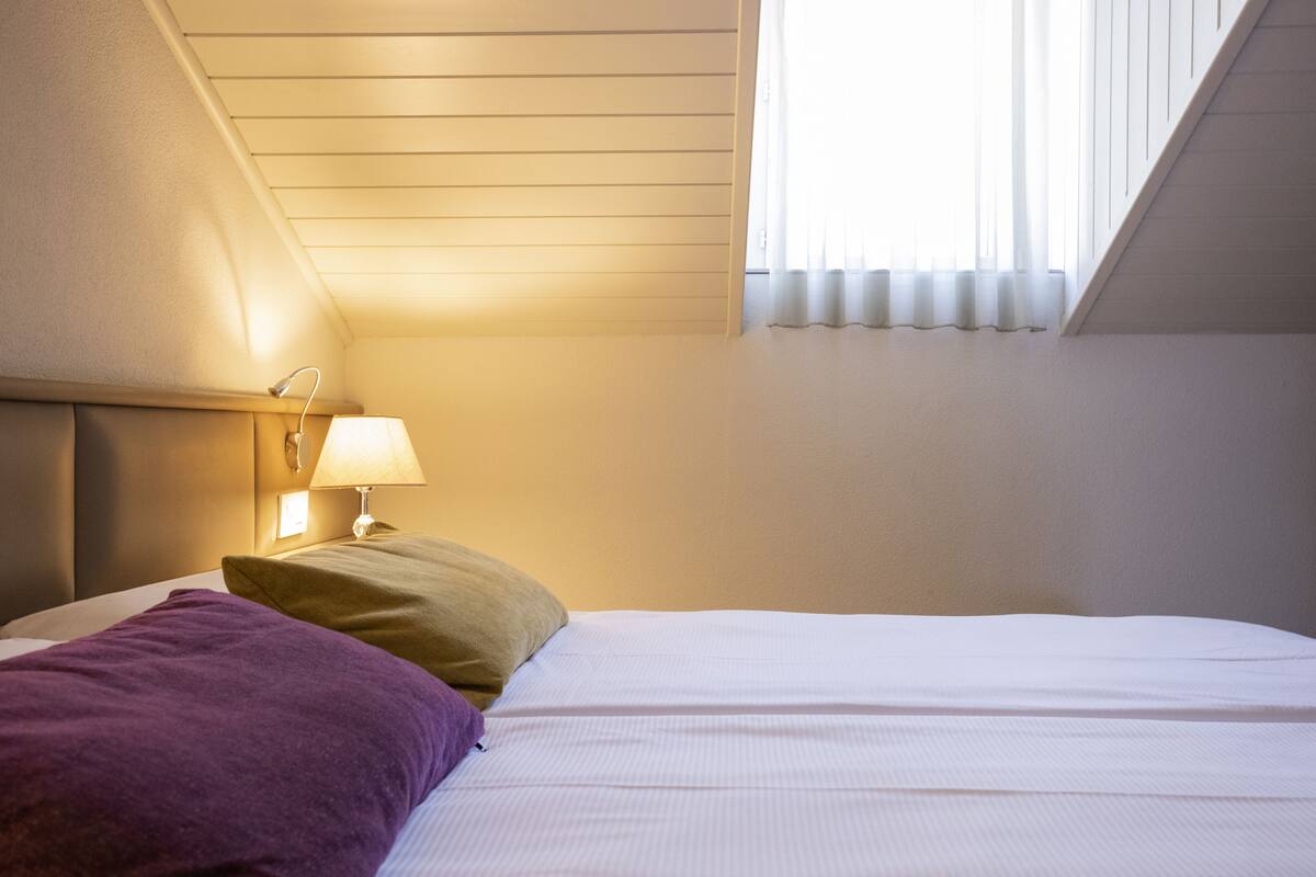 Quarto de hotel com cama, mesa de cabeceira, luminária e janela com cortinas.