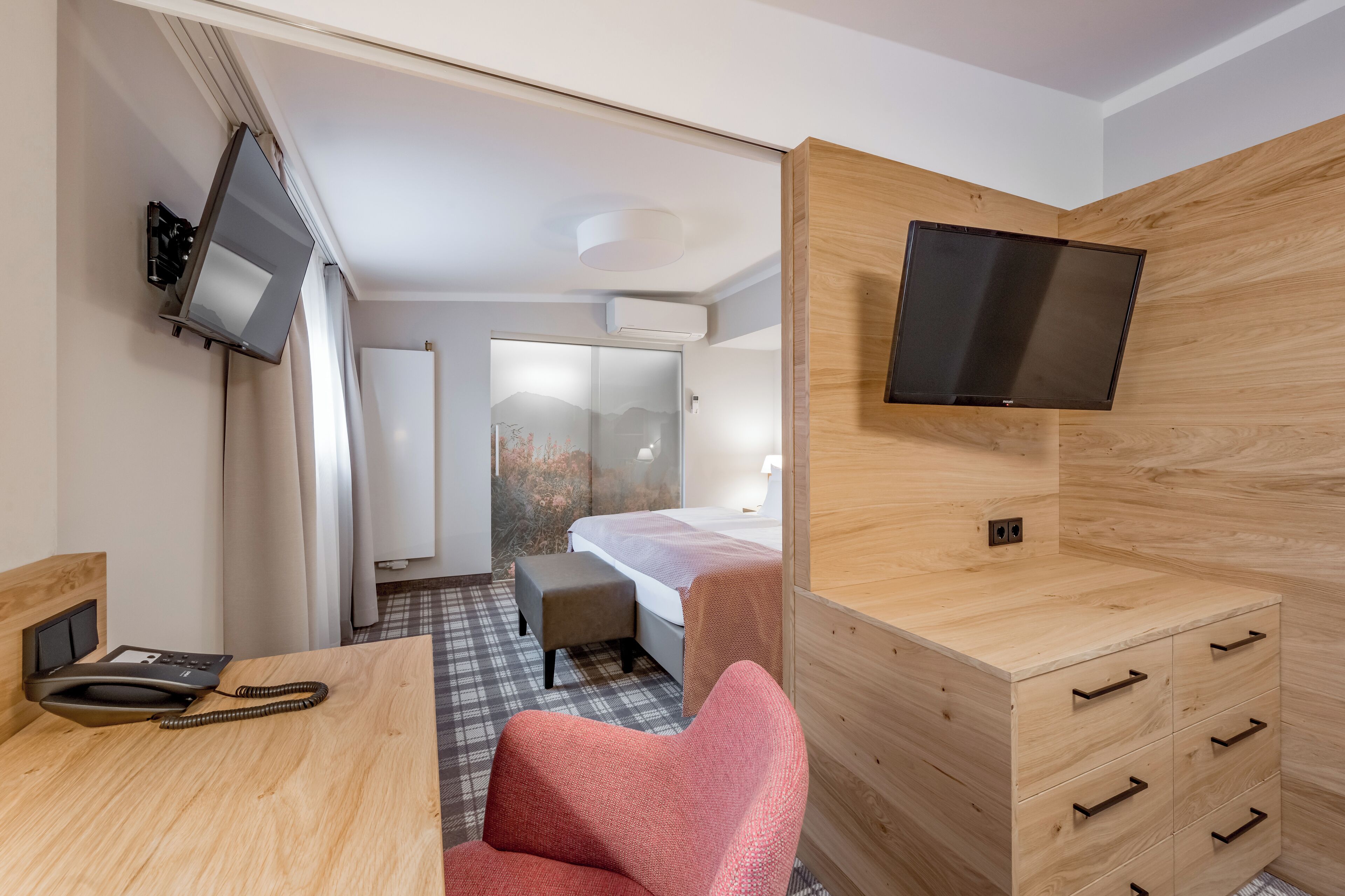 Quarto de hotel com cama, escrivaninha com telefone, televisão fixada na parede e cómoda de madeira.
