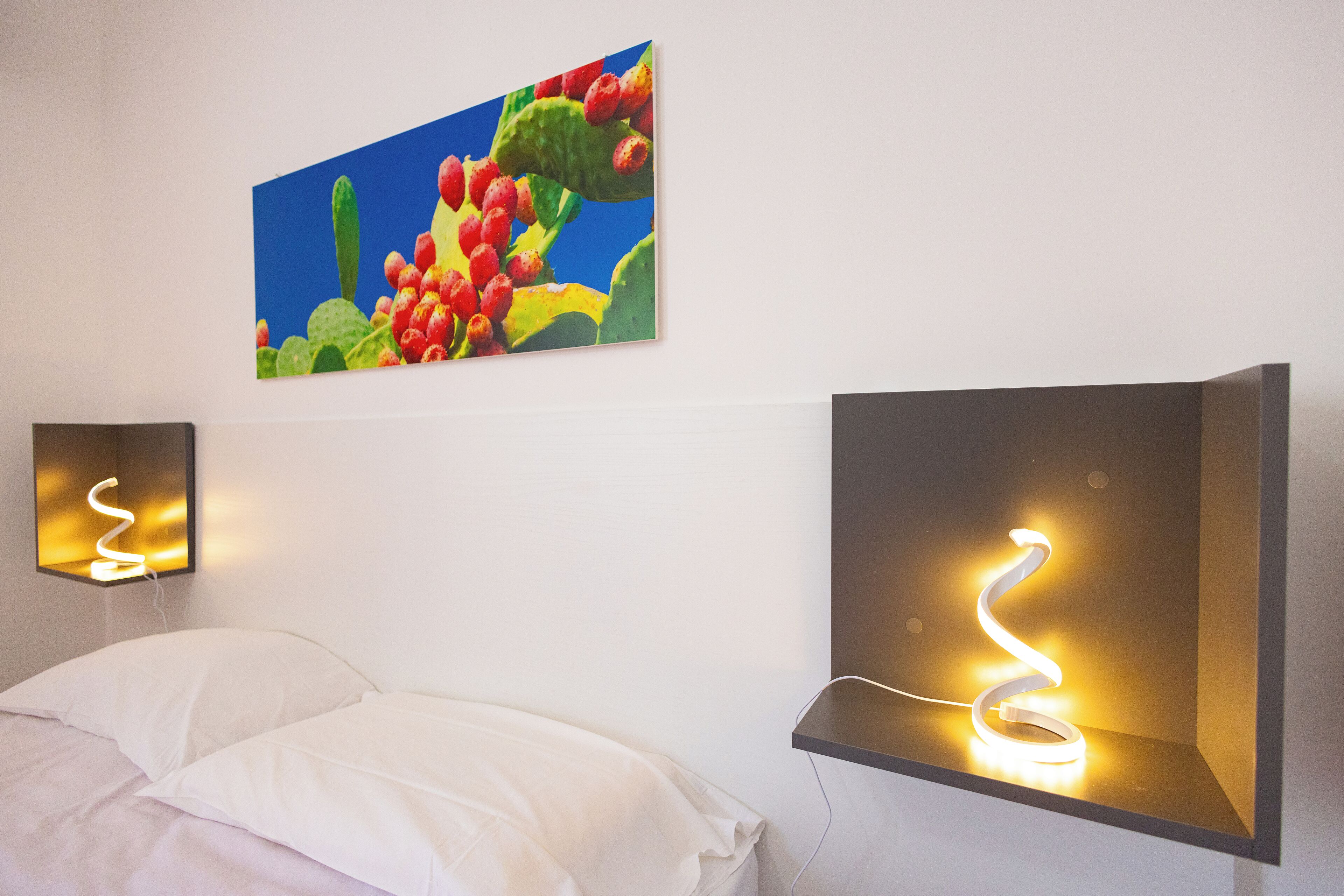 Quarto com uma cama, pintura de cactos pendurada na parede e duas luminárias de parede com design de luz em espiral.