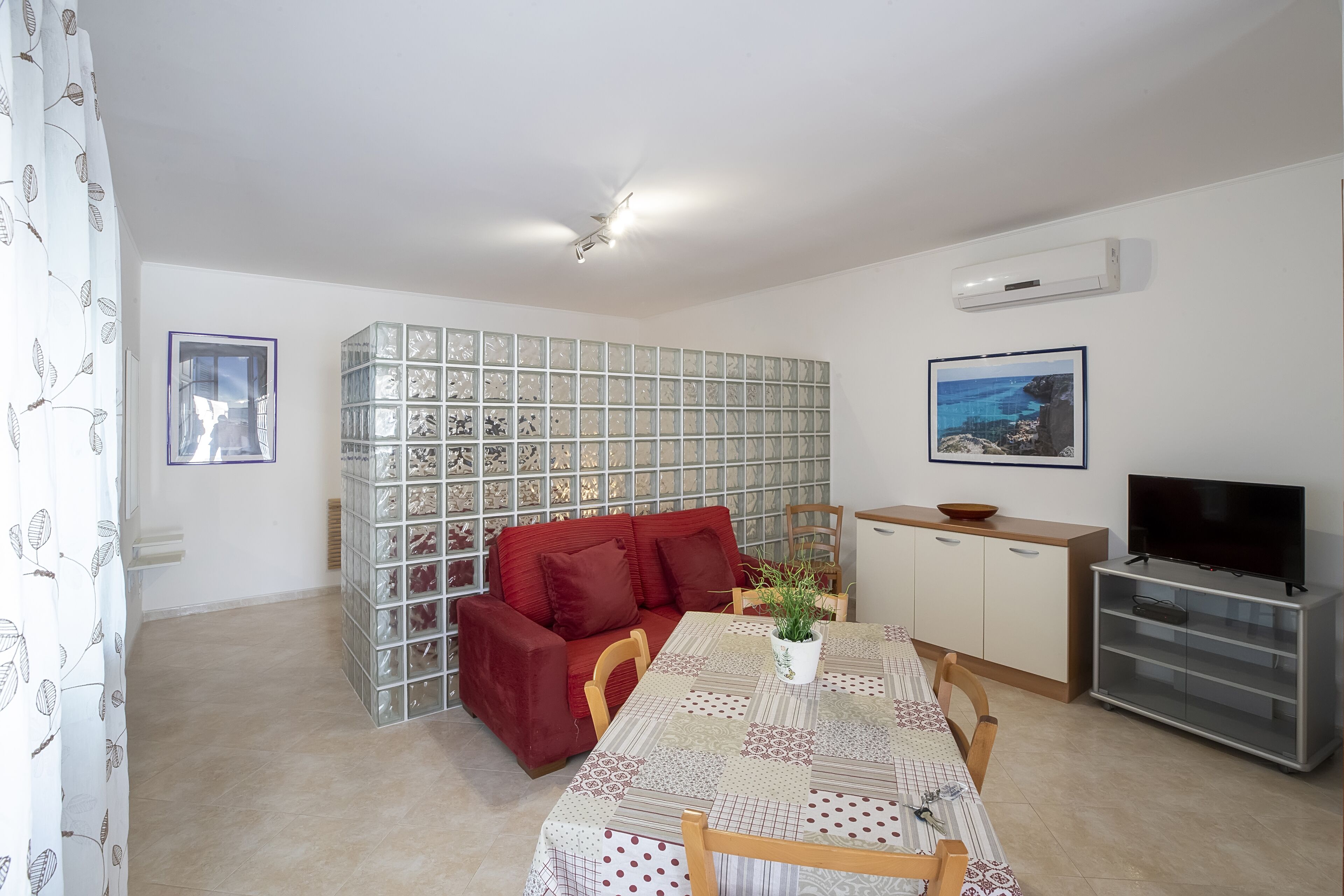 Uma sala de estar com um sofá vermelho, uma mesa de jantar com toalha de mesa estampada, um rack para televisão e um painel de vidro com detalhes decorativos.