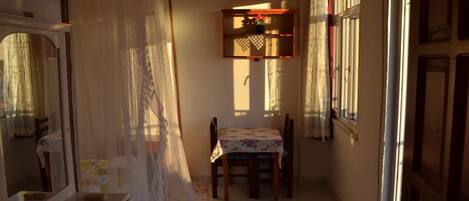 Quarto estreito com teto de madeira, uma mesa com toalha florida, uma cadeira, uma pequena prateleira com uma planta e uma janela com cortinas.