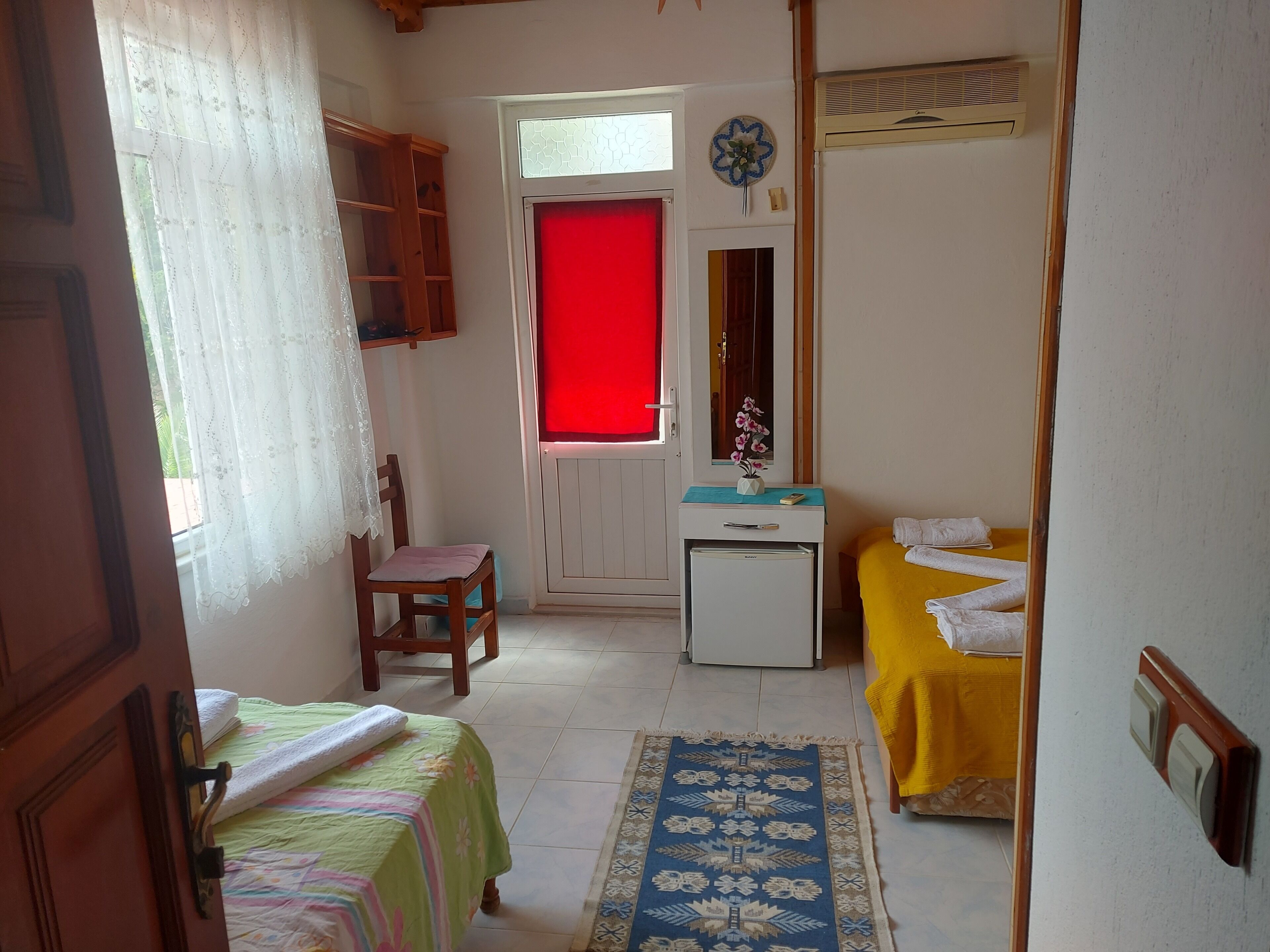 Quarto com uma cama, escrivaninha, cadeira, pequeno frigorífico, espelho e porta com cortina vermelha.