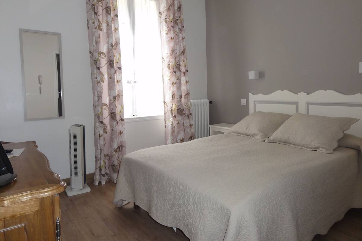 Quarto com cama, cômoda de madeira, radiador e janela com cortinas florais.