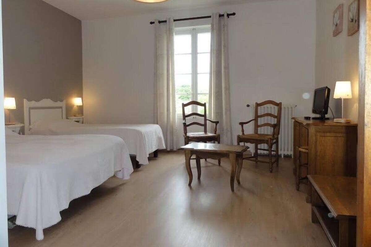 Quarto de hotel com duas camas, uma mesa de madeira, duas cadeiras, uma escrivaninha com um computador e uma janela com cortinas.