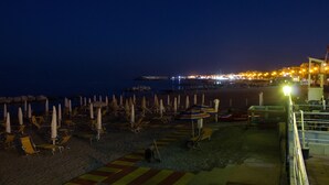 Uma área à beira-mar com fileiras de cadeiras e guarda-sóis de praia, um cais e um edifício bem iluminado ao fundo.