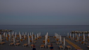 Uma praia com fileiras de espreguiçadeiras e guarda-sóis, um mar calmo e um litoral rochoso.