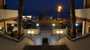 Vista noturna da entrada de um hotel com escadaria, palmeiras e uma rua com carros e um edifício ao fundo.