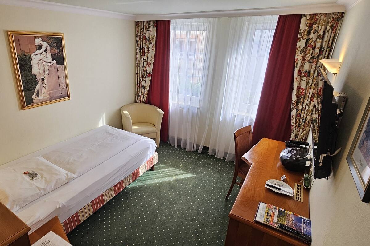 Quarto de hotel com uma cama de solteiro, uma escrivaninha com telefone e livros, uma cadeira, uma janela com cortinas e um quadro na parede.