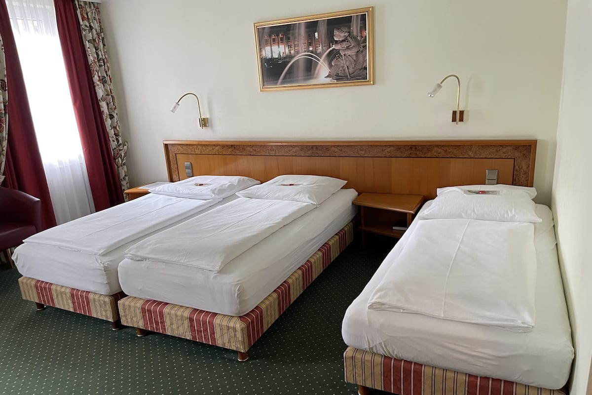 Quarto de hotel com duas camas de solteiro, cabeceira de madeira, mesinhas de cabeceira pequenas de madeira, um quadro na parede e um tapete verde com padrão de bolinhas.