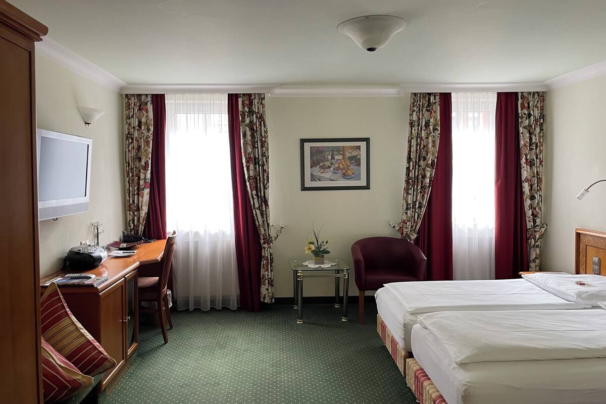 Quarto de hotel com cama, escrivaninha, cadeira, televisão e uma pequena mesa com um vaso.