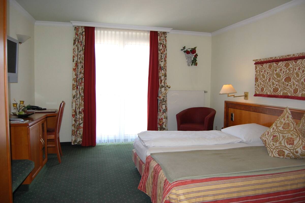 Quarto de hotel com duas camas, uma escrivaninha, uma cadeira, um abajur e uma janela com cortinas.
