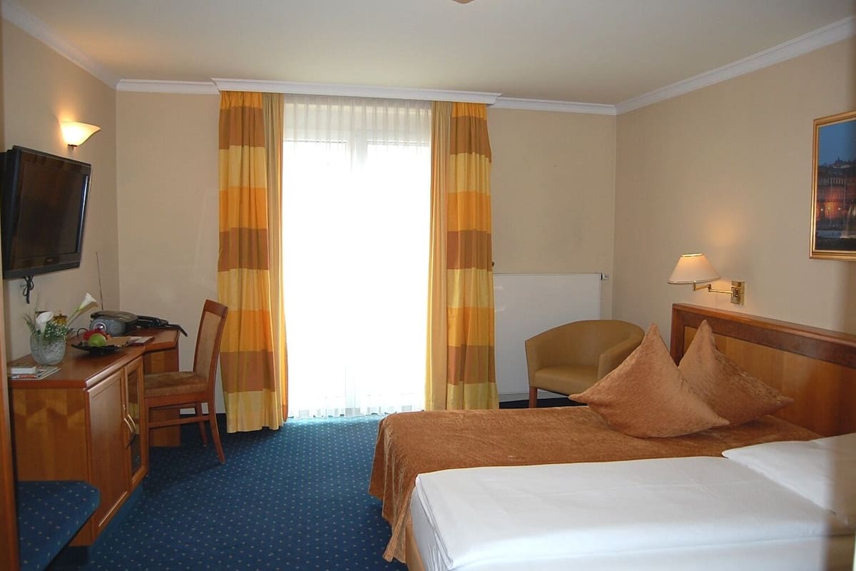 Quarto de hotel com uma cama, uma escrivaninha com cadeira, televisão e uma janela com cortinas.
