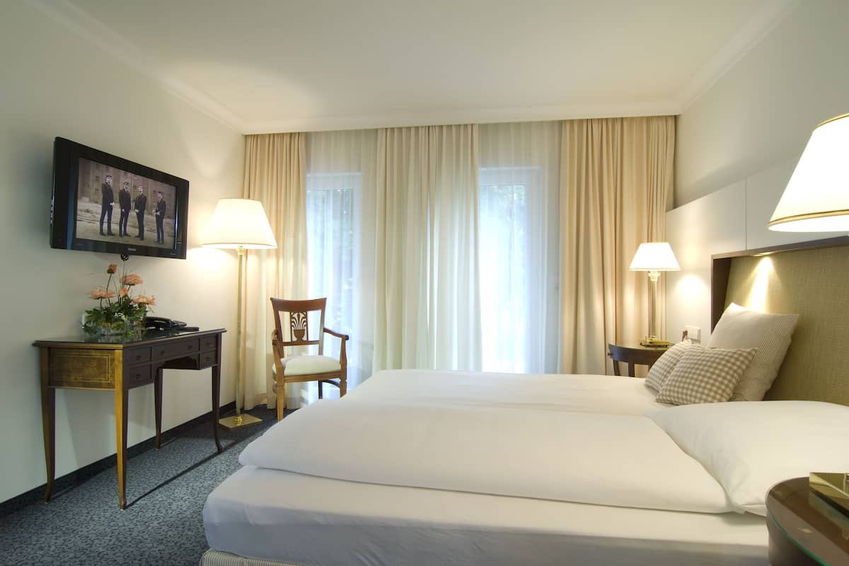 Quarto de hotel com uma cama grande, uma escrivaninha com abajur, uma cadeira e televisão na parede.