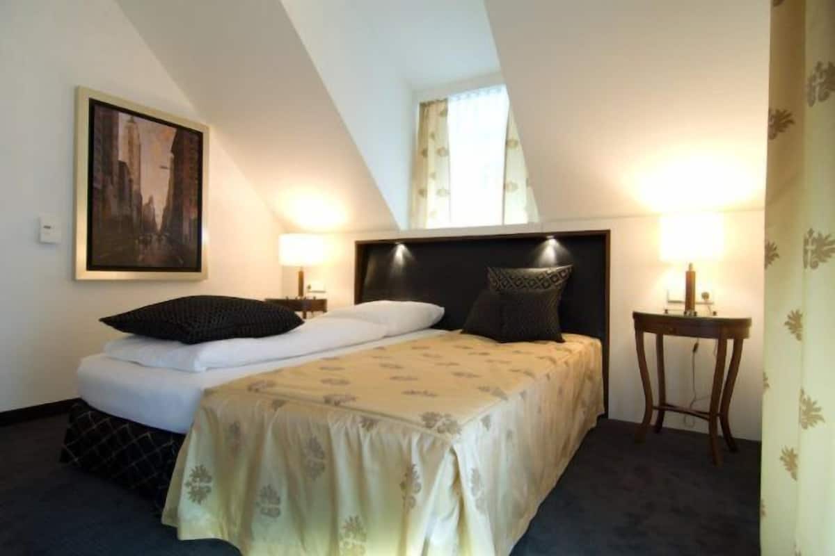 Quarto de hotel com uma cama, duas luminárias de cabeceira, um quadro na parede e uma janela com cortinas.