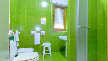 Casa de banho com azulejos verdes, sanita, lavatório, chuveiro e um pequeno banquinho.