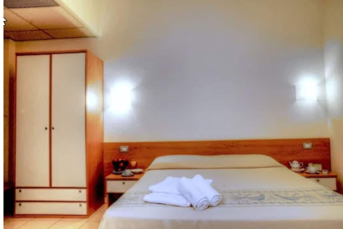 Quarto de hotel com cama, cabeceira de madeira, mesa de cabeceira e guarda-roupa.